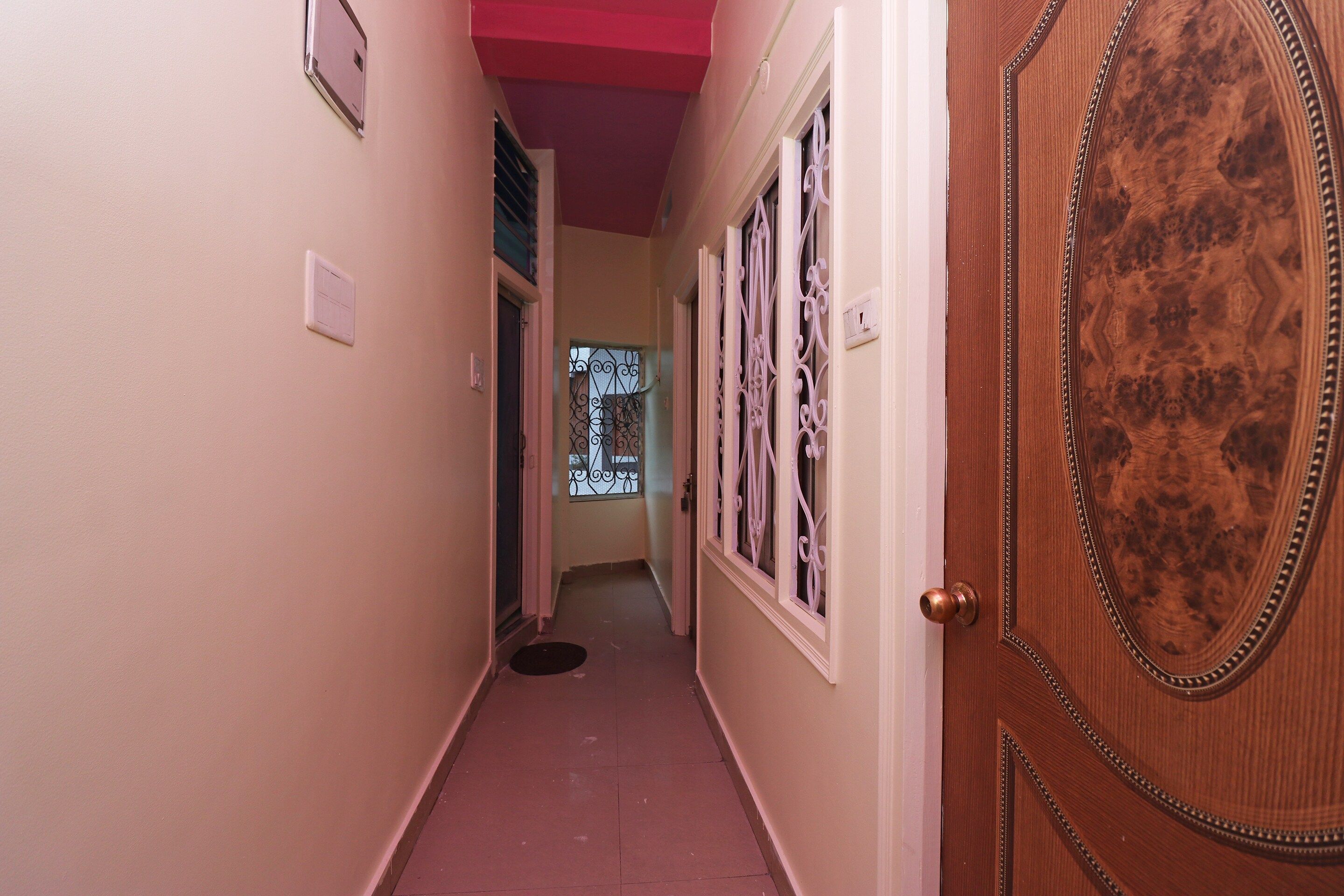 Um corredor estreito com uma porta de madeira à direita, uma janela com grades de ferro decorativas e um interfone à esquerda.