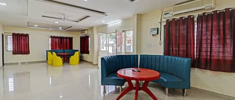 Quarto de hotel com sofá azul, mesa vermelha, cadeiras amarelas e cortina vermelha.