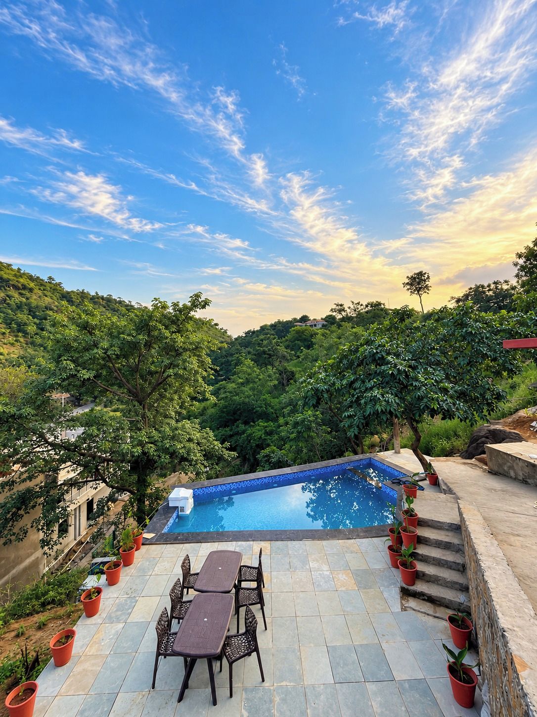 Piscina com deck revestido, rodeada por vasos de plantas e mesa e cadeiras para refeições ao ar livre.