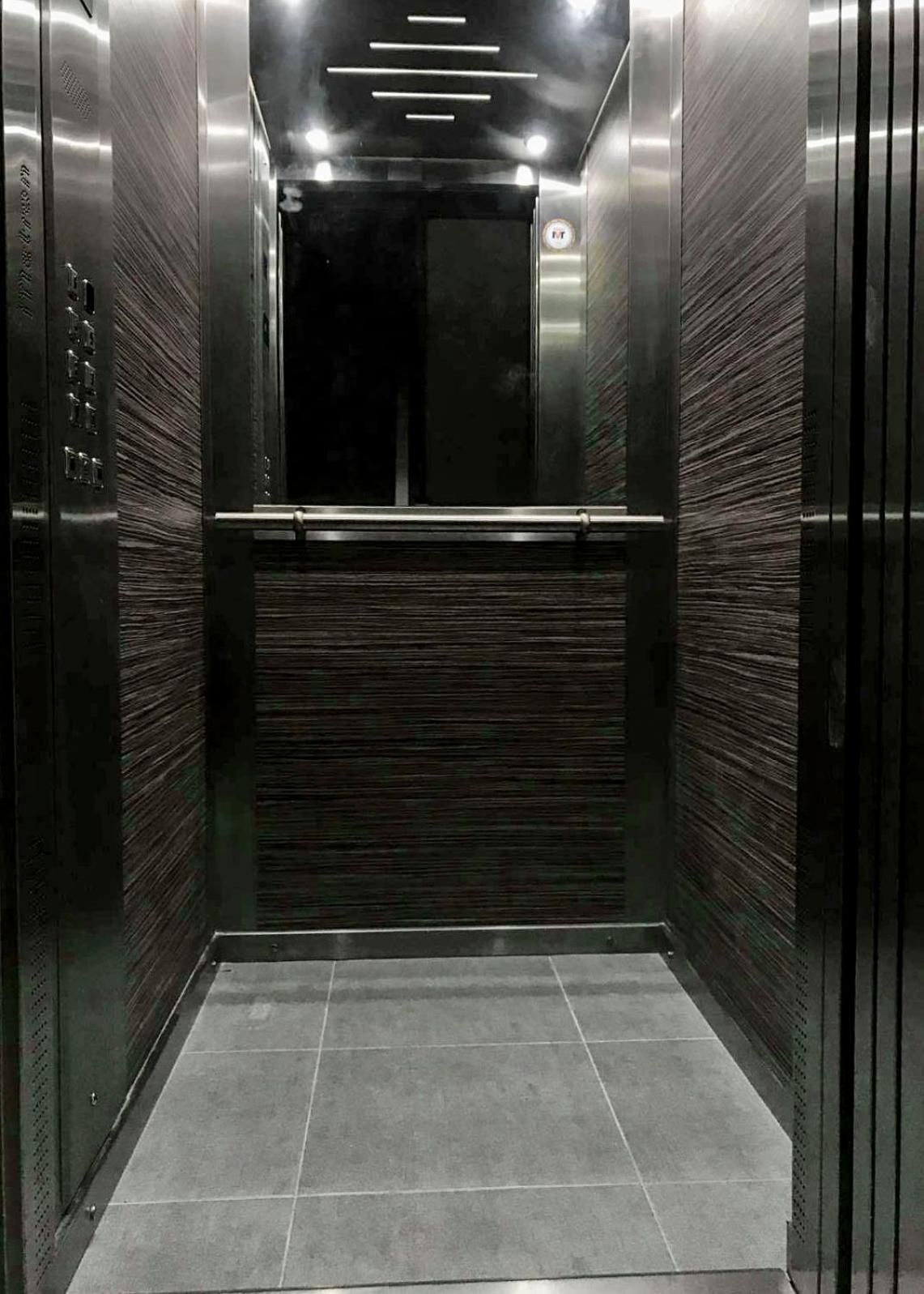Interior de elevador com paredes metálicas, piso ladrilhado e painel de controle à esquerda.