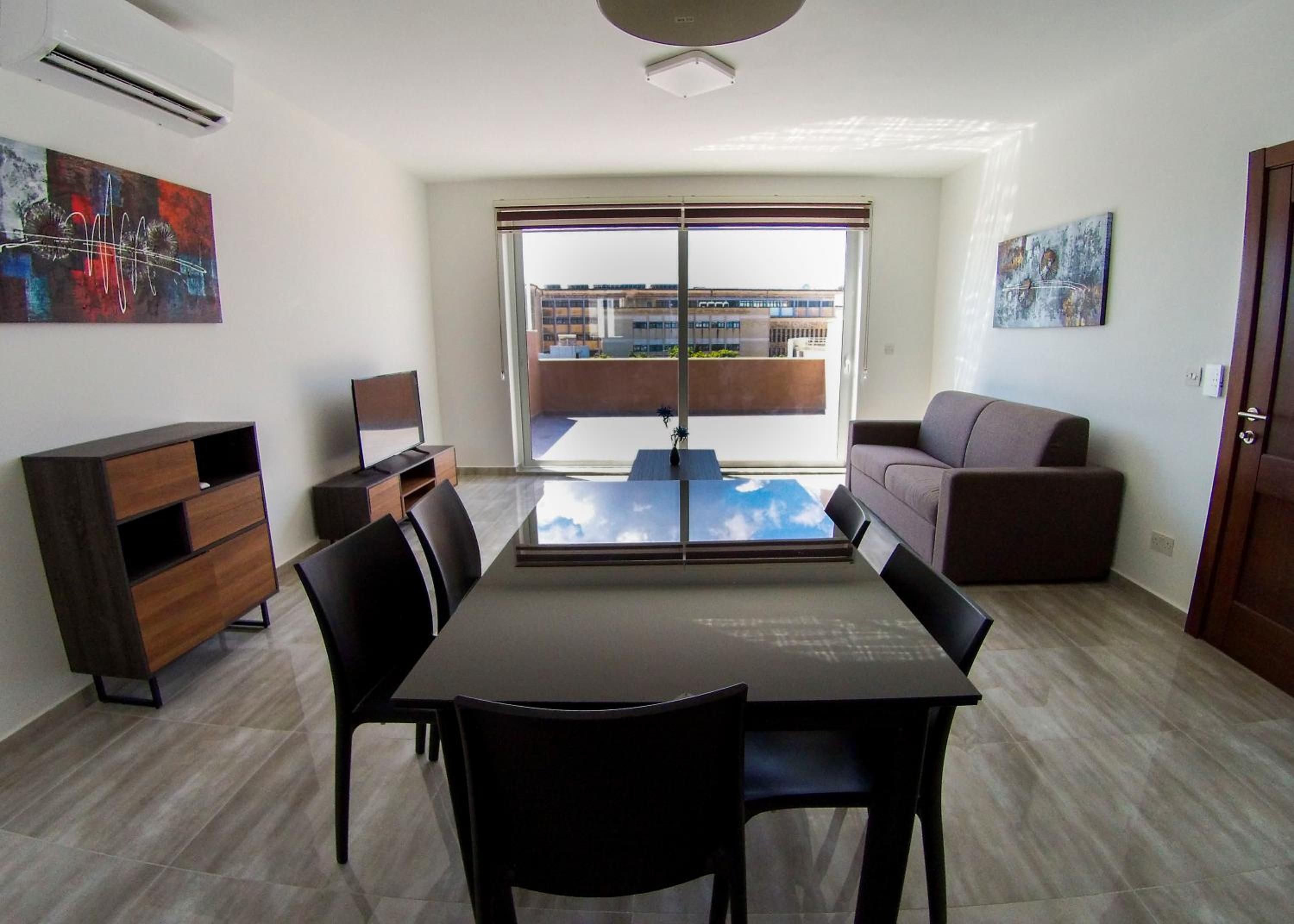 Uma sala de estar moderna com uma mesa de jantar, cadeiras, um sofá e uma grande janela com vista para um edifício.