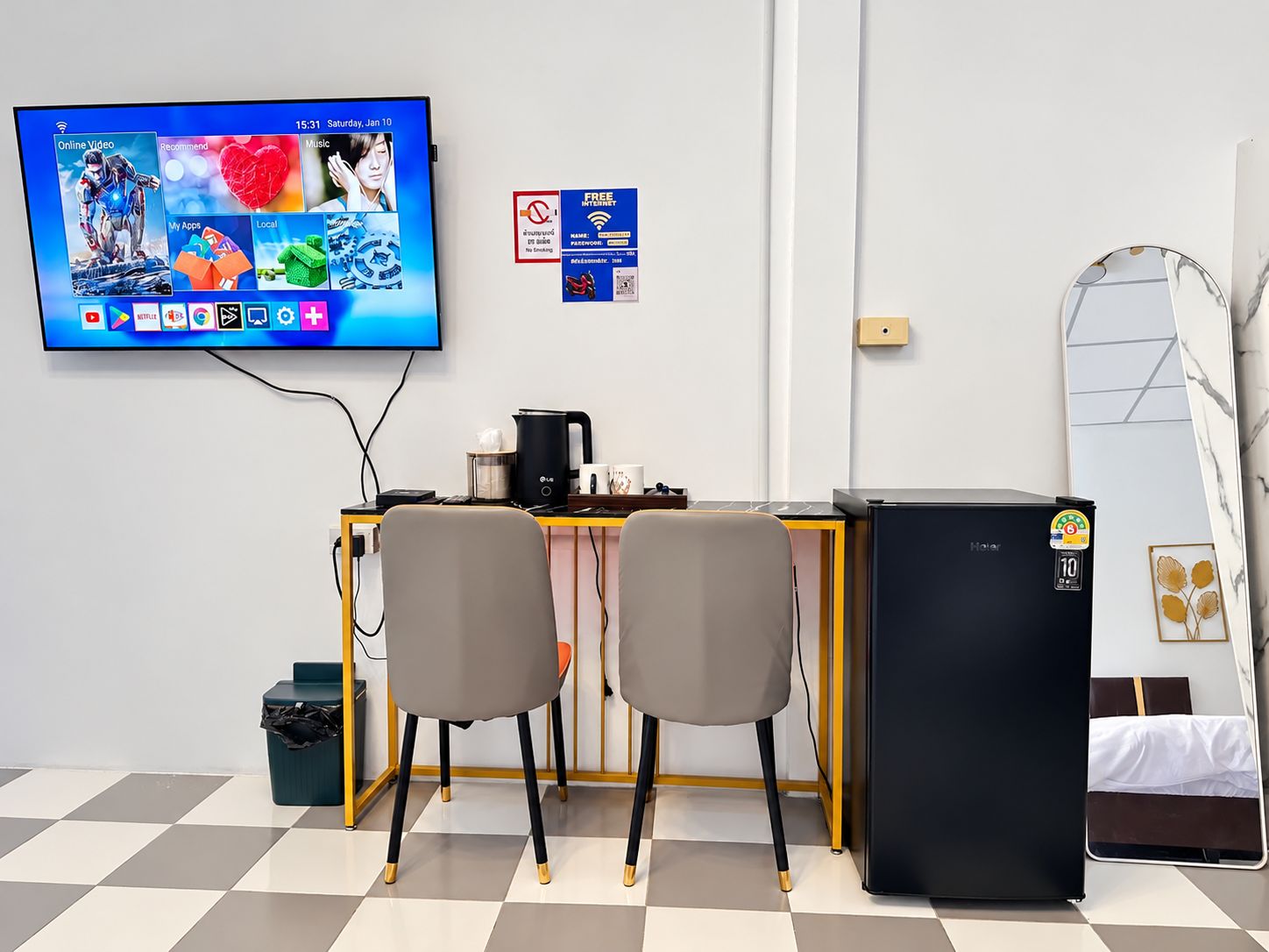 Quarto de hotel com escrivaninha, duas cadeiras, televisão exibindo diversos aplicativos, cafeteira e geladeira.