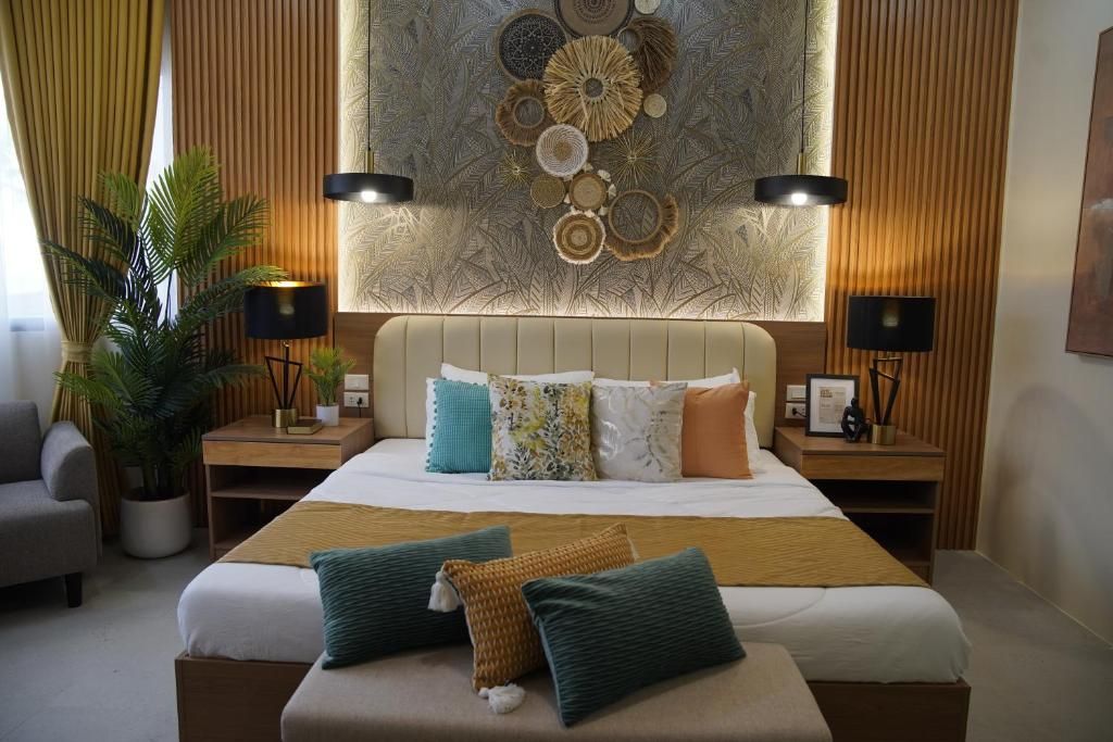Quarto de hotel com uma cama grande, duas mesas de cabeceira, um vaso de plantas e um objeto decorativo na parede.