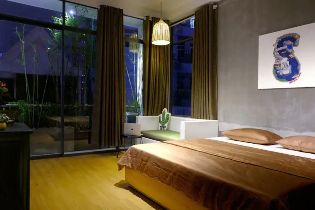 Quarto com cama, escrivaninha, cadeira e uma janela grande com cortinas.