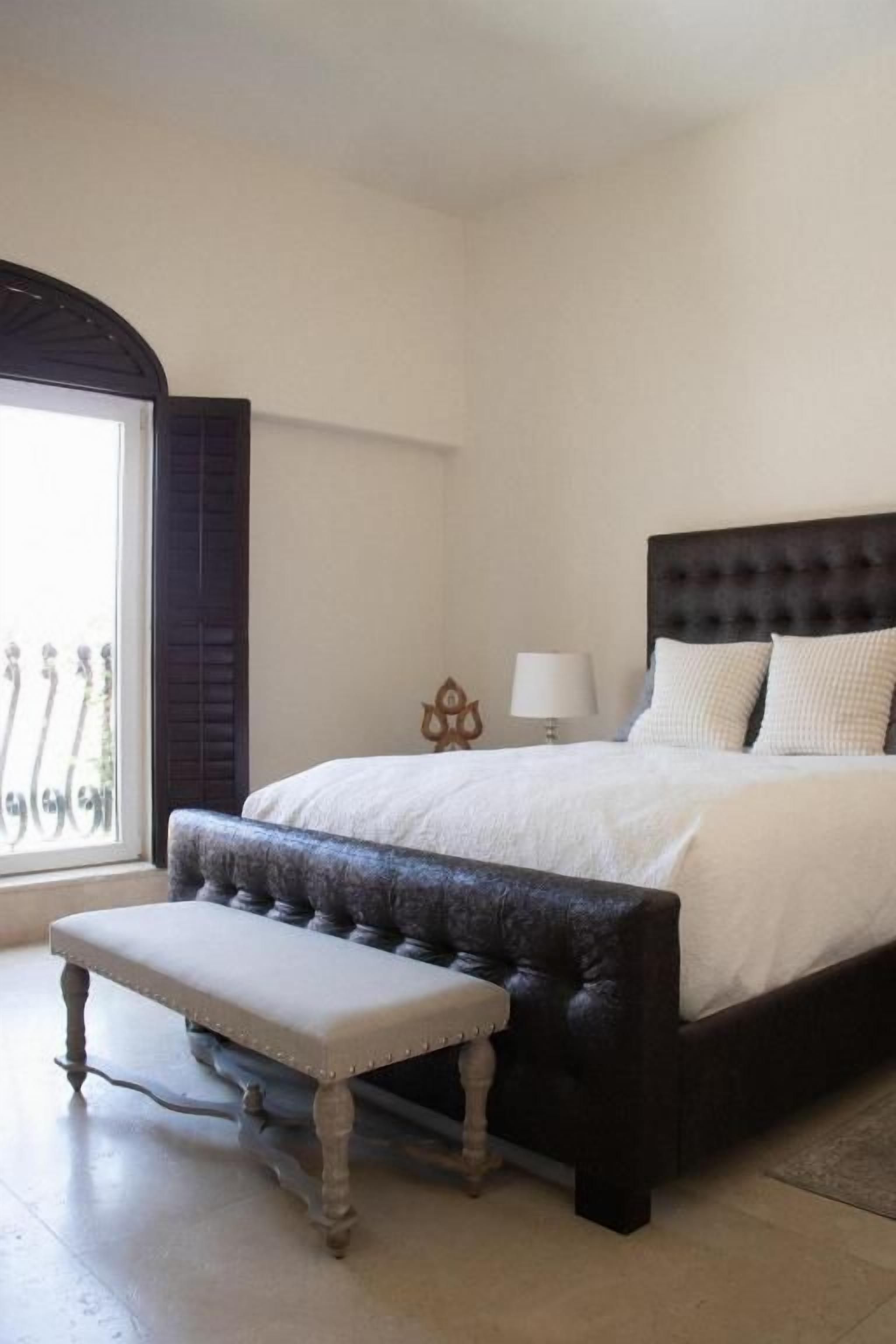 Quarto com cama grande, um banco, mesa de cabeceira e janela.