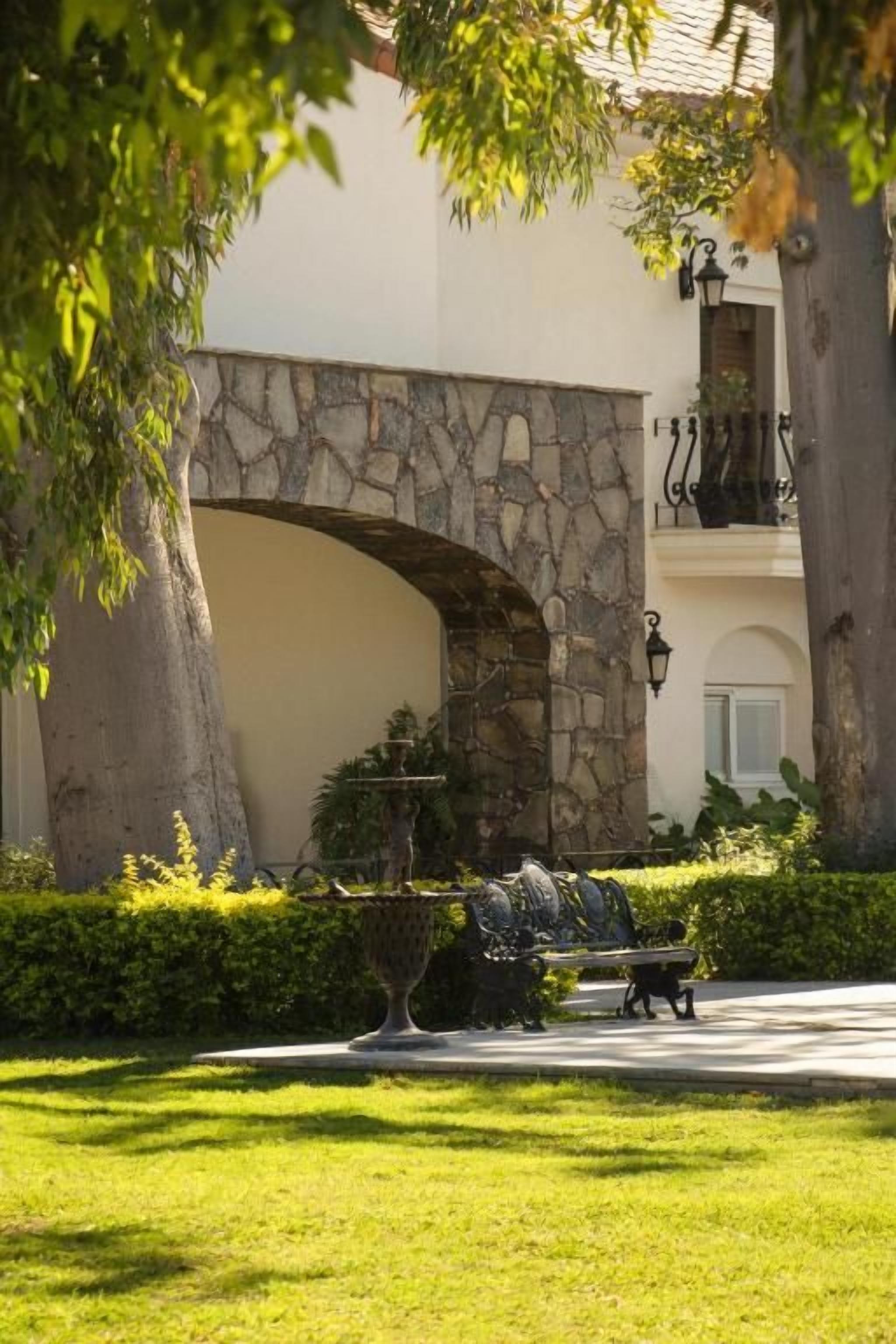Arcada de pedra que dá acesso a um edifício, uma fonte e um jardim bem cuidado.