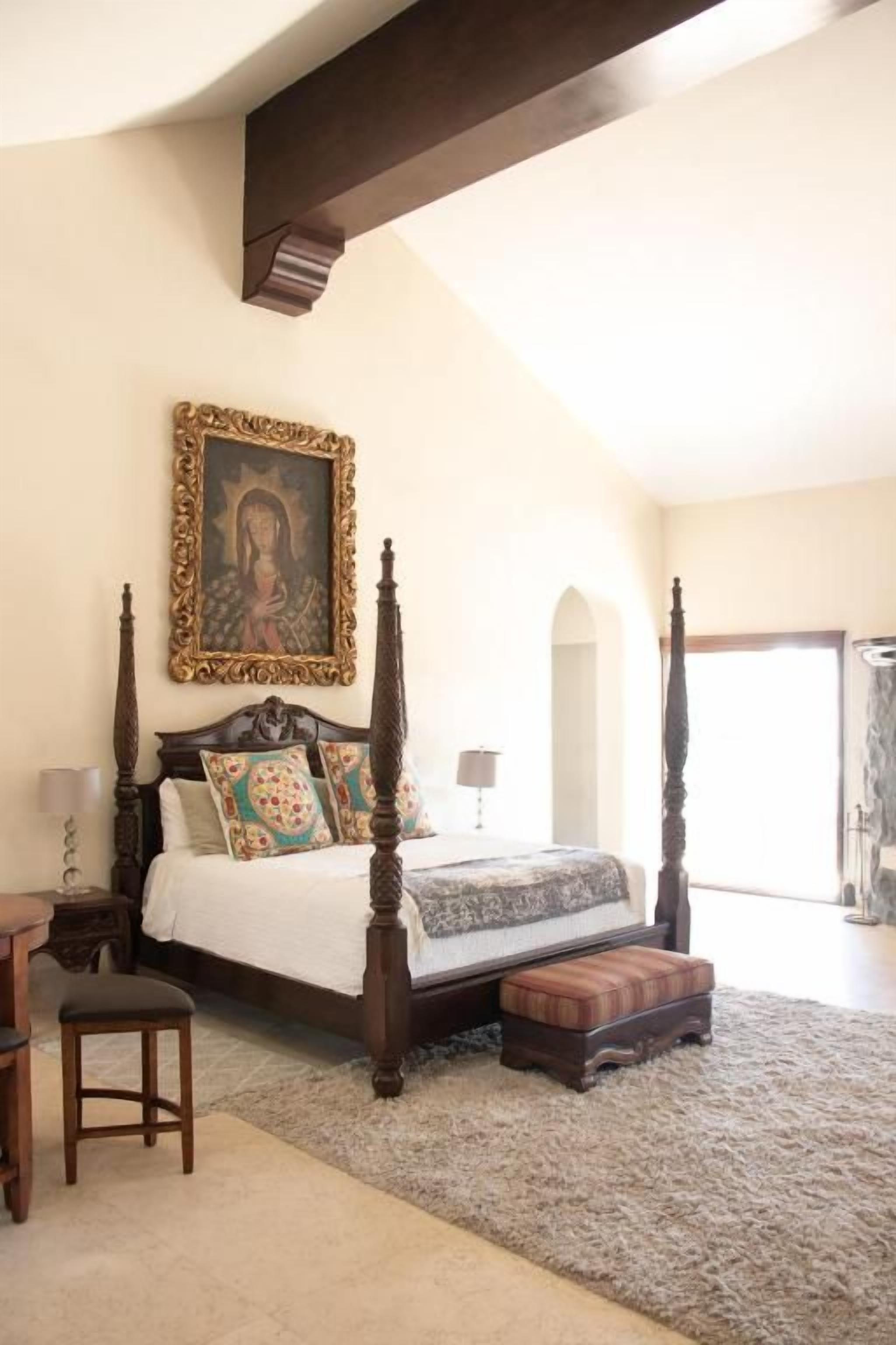 Quarto com cama de dossel, mesa de cabeceira, uma cadeira e uma grande moldura na parede.