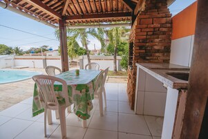 Um pátio ao ar livre com piscina, área de refeições coberta com mesa e cadeiras, e uma parede de tijolos.