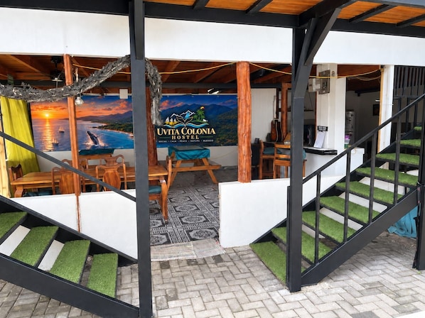 Interior de um hostel com um mural, móveis de madeira e uma escada coberta com grama sintética verde.