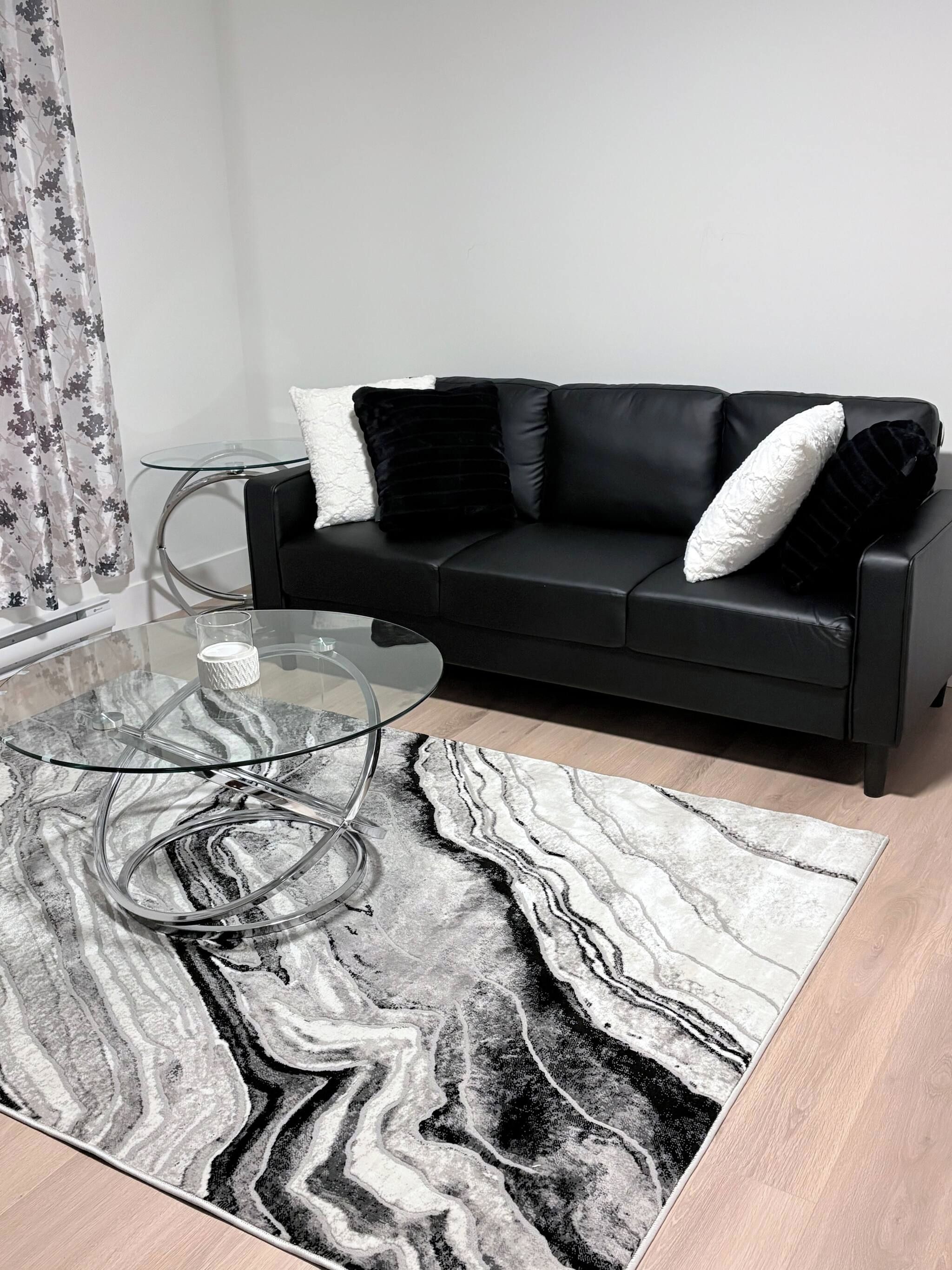 Uma sala de estar moderna com uma mesa de centro de vidro, um sofá de couro preto com almofadas estampadas e um tapete com padrão marmorizado.