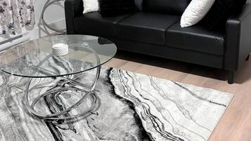 Uma sala de estar moderna com uma mesa de centro de vidro, um sofá de couro preto com almofadas estampadas e um tapete com padrão marmorizado.
