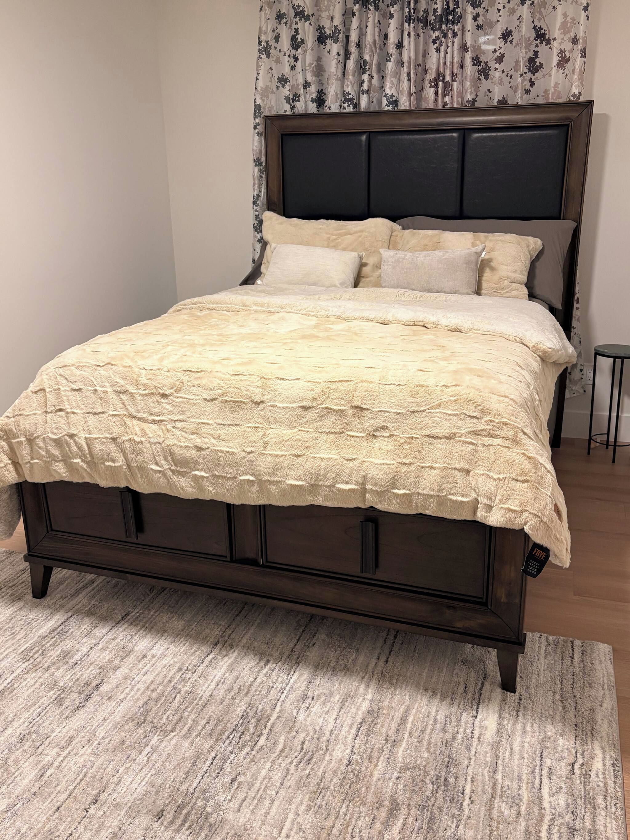 Uma cama com estrutura de madeira escura, colcha bege e cabeceira combinando.