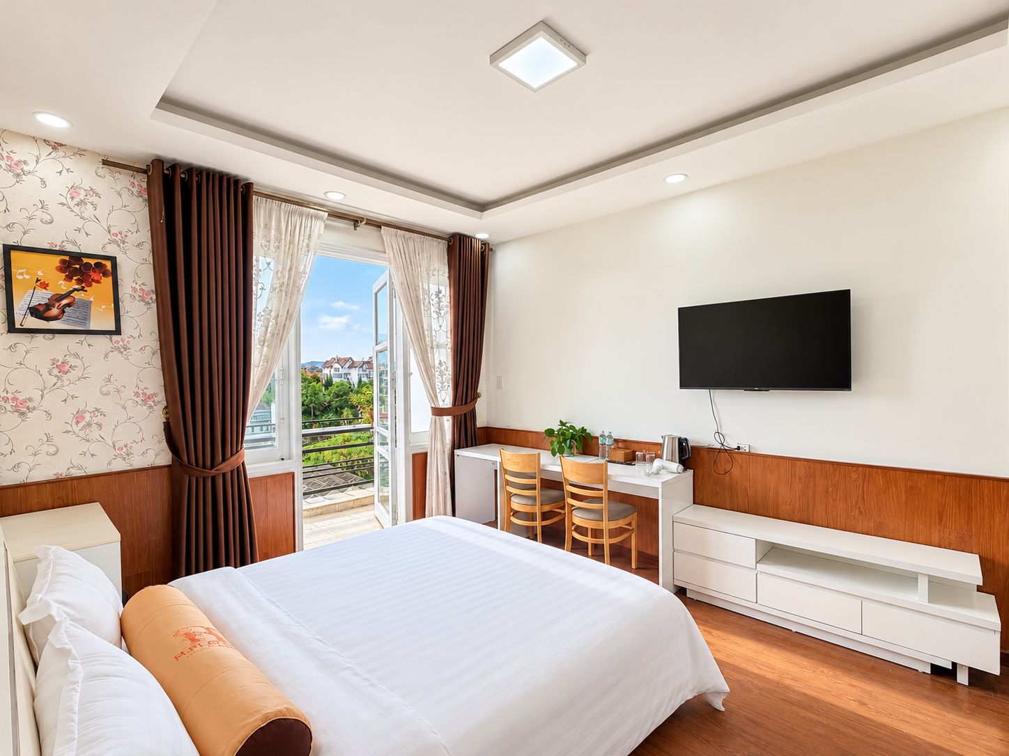 Quarto de hotel com cama, escrivaninha com cadeiras, televisão e varanda com cortinas.