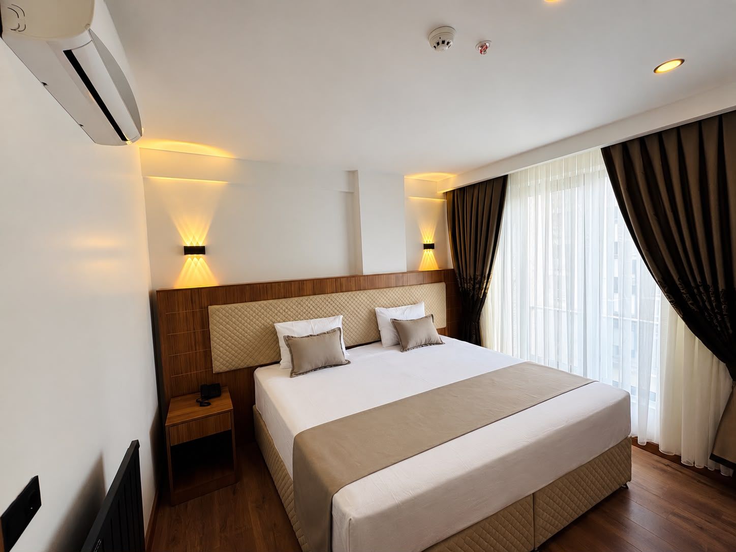 Quarto de hotel com uma cama grande, mesas de cabeceira, aparelho de ar condicionado, luminárias fixadas na parede e uma janela com cortinas.