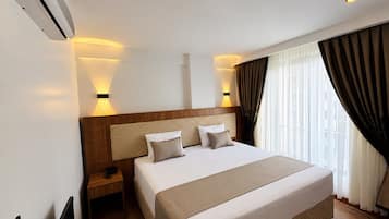 Quarto de hotel com uma cama grande, mesas de cabeceira, aparelho de ar condicionado, luminárias fixadas na parede e uma janela com cortinas.