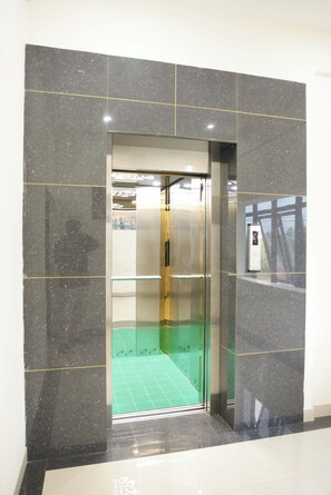 Um elevador com tapete de segurança verde na entrada, rodeado por azulejos escuros com manchas.