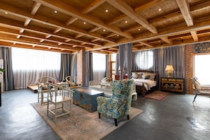 Uma sala de estar ampla, com teto de madeira alto, uma cama grande e um espaço de estar com uma poltrona estampada.