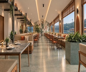 Um restaurante com um amplo espaço de refeições, várias mesas e cadeiras, e vista para um lago e uma floresta ao fundo.
