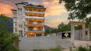 Um hotel com vários andares, varandas, uma placa com o texto 'The Ravasa' e montanhas ao fundo.