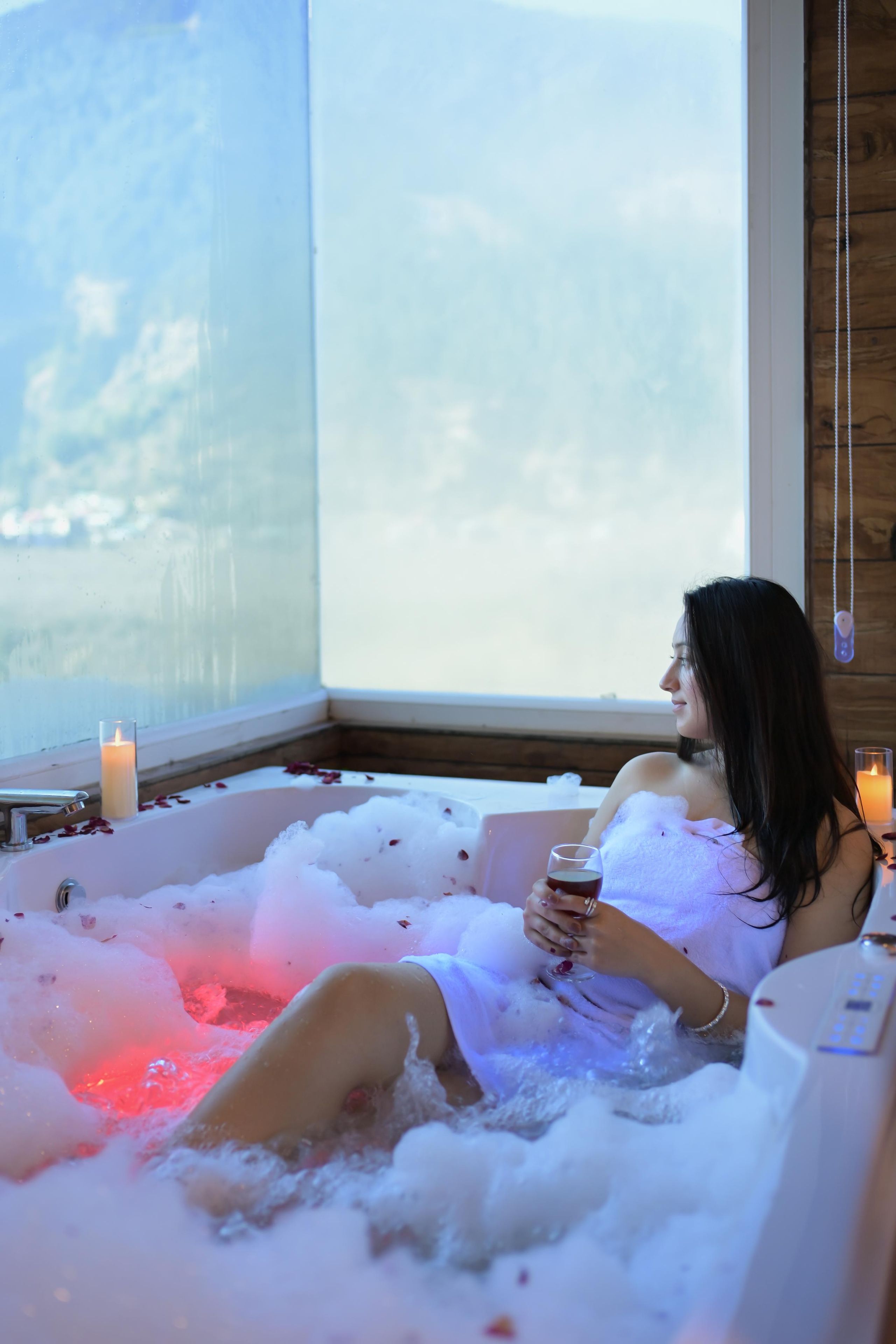 Uma pessoa relaxando em uma banheira cheia de espuma, segurando um copo de bebida, com velas e vista pela janela.