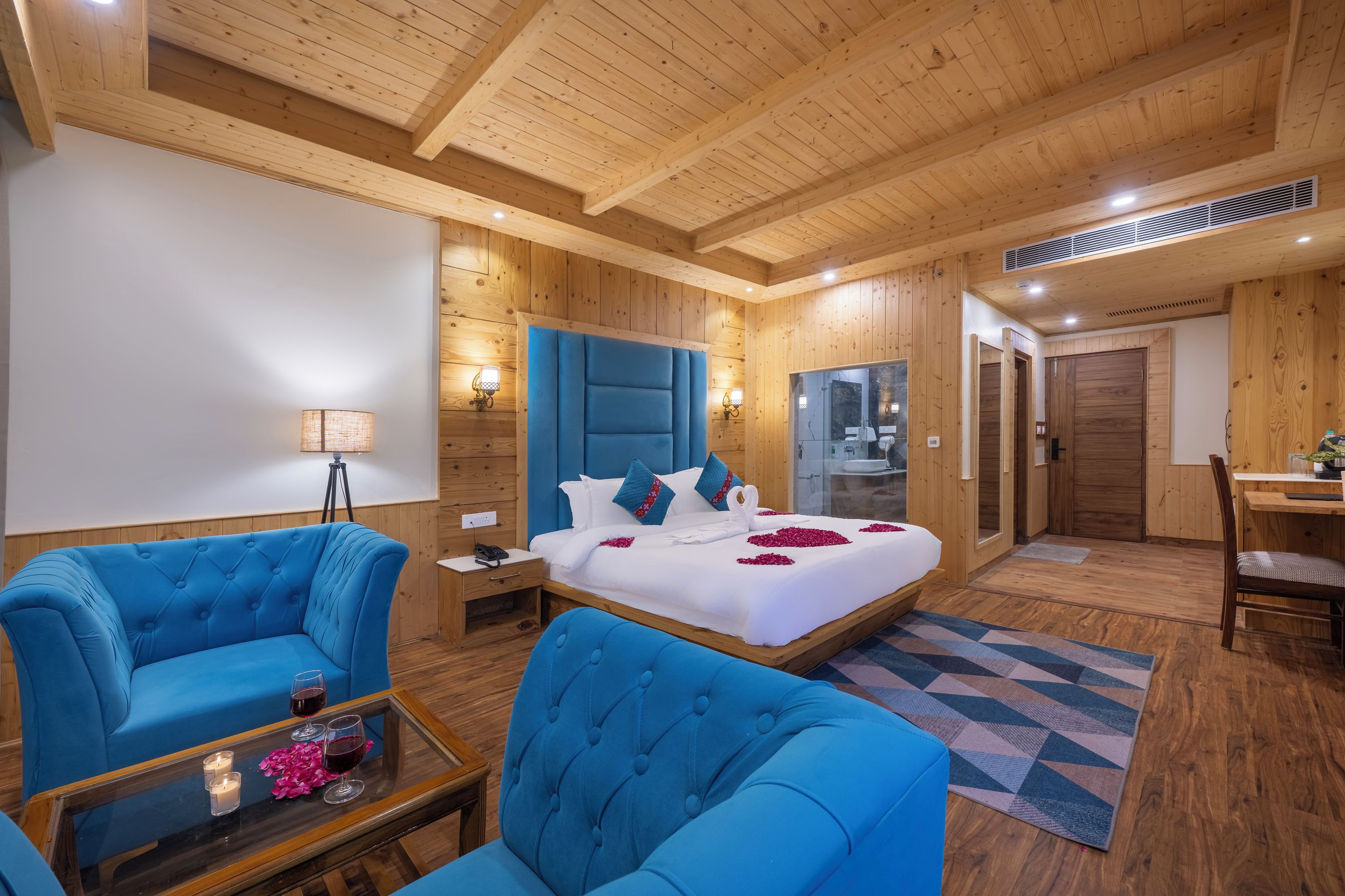 Quarto de hotel com teto de madeira, uma cama grande com cabeceira azul, duas poltronas azuis, uma pequena mesa com uma vela e copos, e um banheiro visível através de uma porta de vidro.