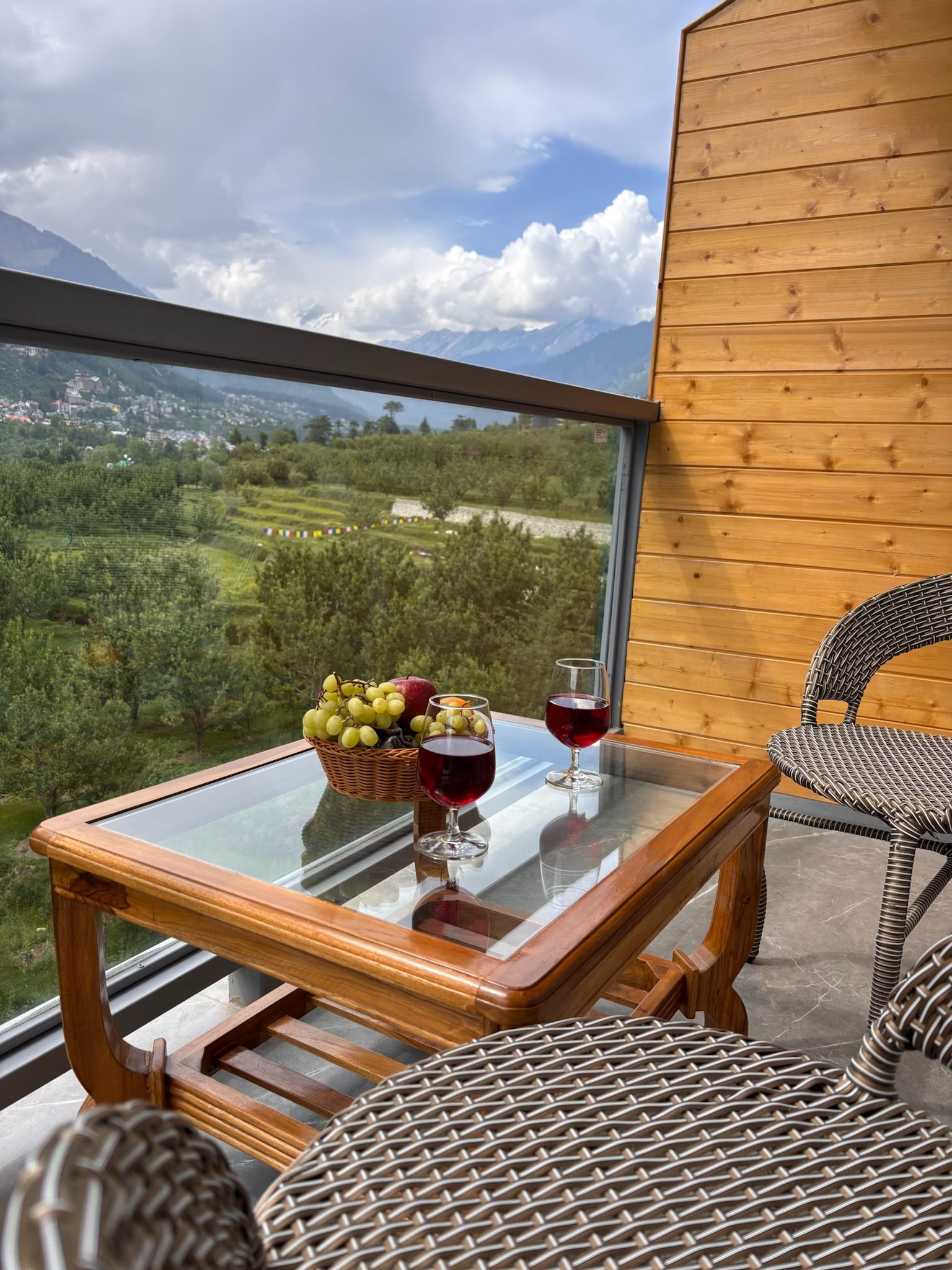 Uma varanda com mesa de vidro, cadeiras de vime e vista para uma paisagem montanhosa.