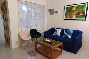 Uma sala de estar com um sofá azul, uma mesa de centro de madeira, uma poltrona rosa, uma poltrona verde e um quadro na parede.