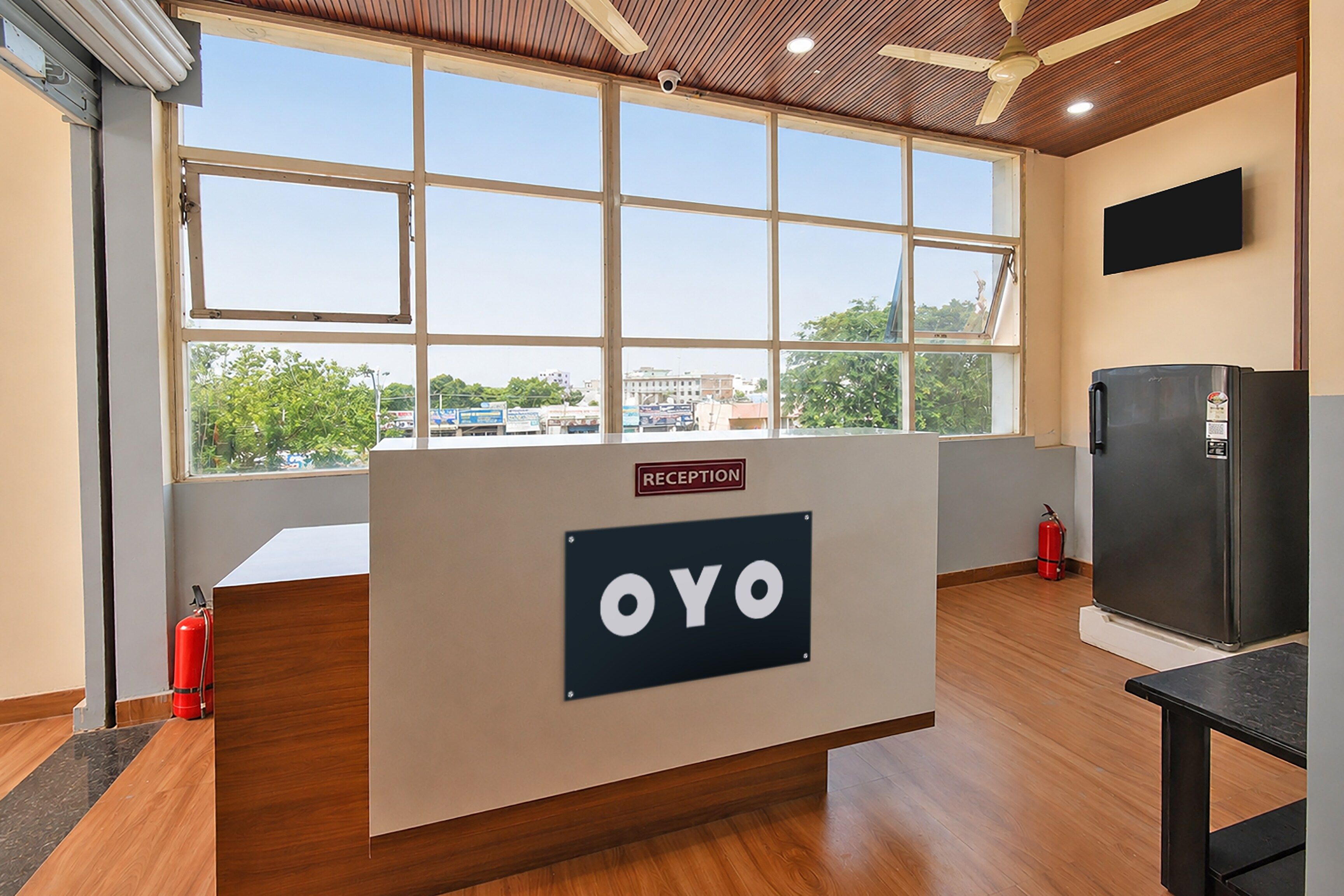 Uma área de recepção com balcão identificado como 'OYO', um extintor de incêndio, uma geladeira e vista para o exterior através de grandes janelas.