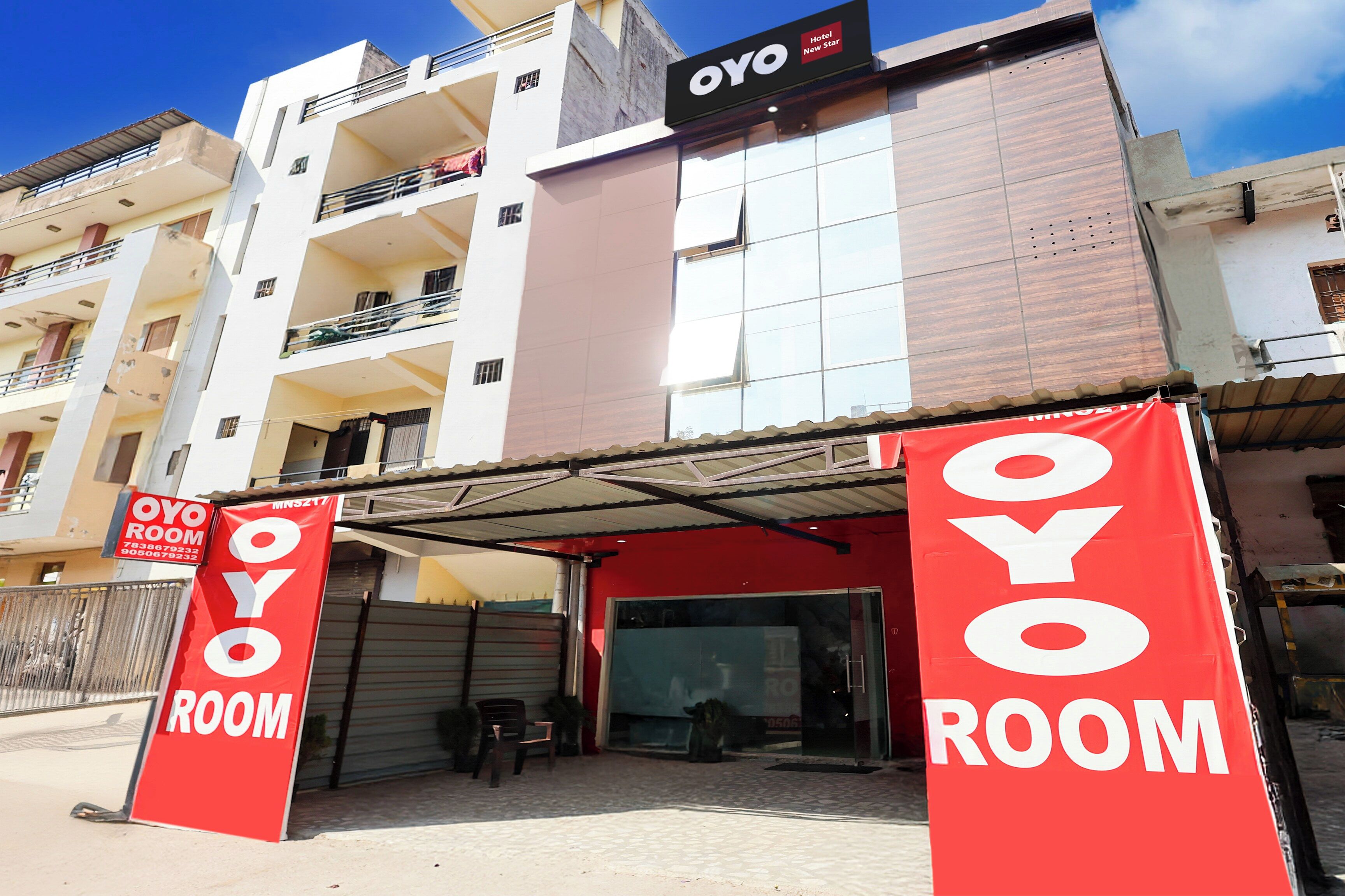 Um hotel chamado OYO, com toldo vermelho e placas, um edifício de vários andares com varandas, e um céu azul claro.