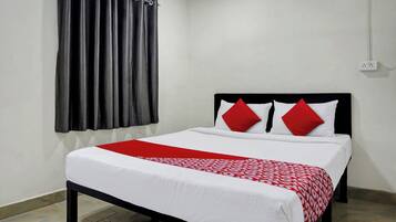 Uma cama de solteiro com colcha branca e vermelha, duas almofadas vermelhas e cabeceira preta em quarto com paredes brancas e janela com cortinas cinzas.