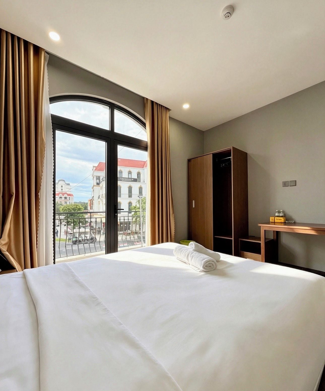 Quarto de hotel com cama grande, guarda-roupa, escrivaninha e vista para edifícios pela janela.