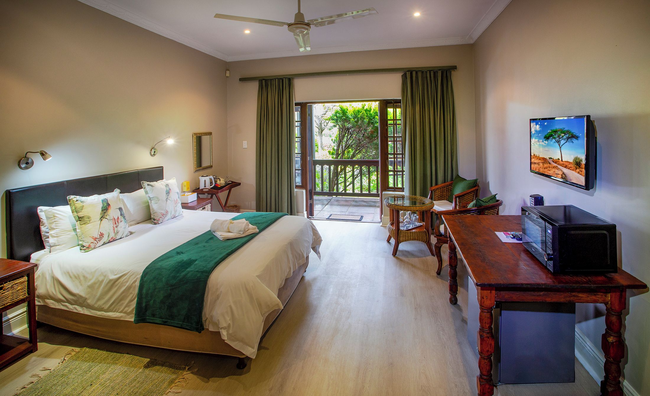 Quarto de hotel com cama, escrivaninha, televisão e varanda com vista para o verde.