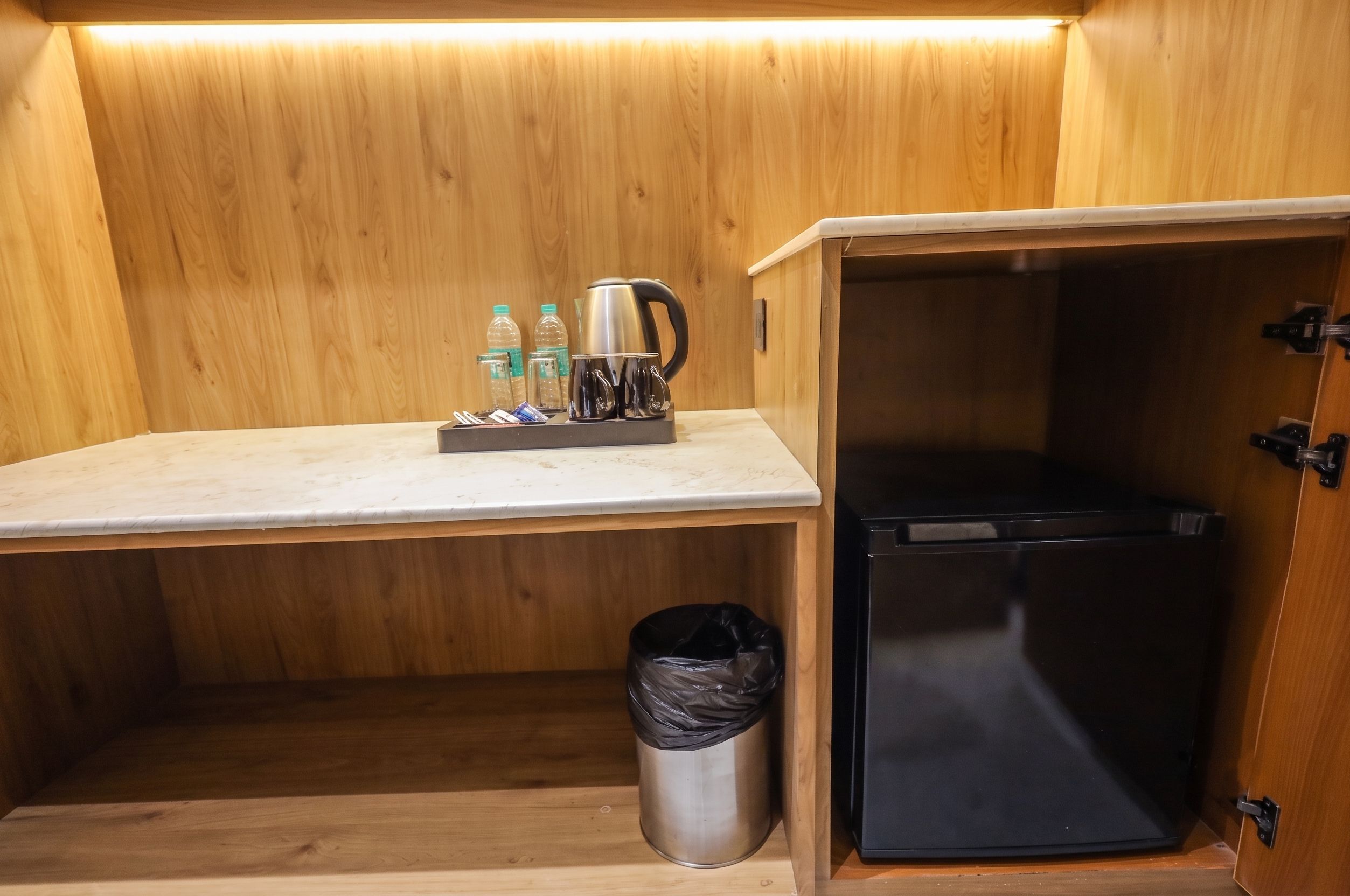 Uma cozinha compacta com bancada, cafeteira, garrafas de água e um cesto de lixo.