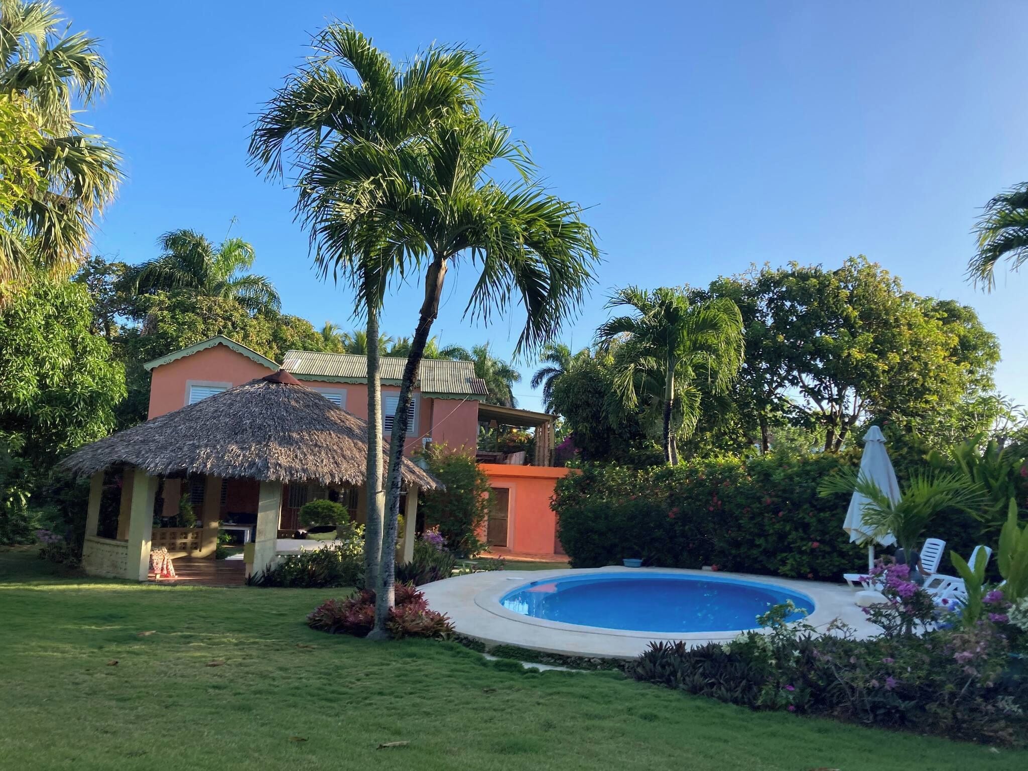 Piscina cercada por um gramado bem cuidado, uma estrutura com telhado de palha e uma casa com fachada vermelha.