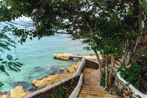 Escadaria descendo até uma orla rochosa com água cristalina, cercada por vegetação exuberante.