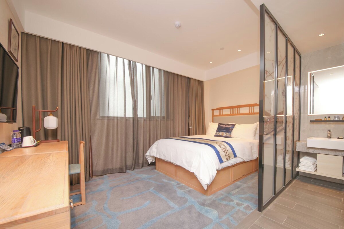 Quarto de hotel com cama, escrivaninha, espelho, banheiro com pia e uma janela grande com cortinas.