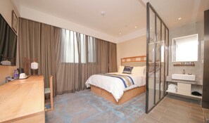 Quarto de hotel com cama, escrivaninha, espelho, banheiro com pia e uma janela grande com cortinas.