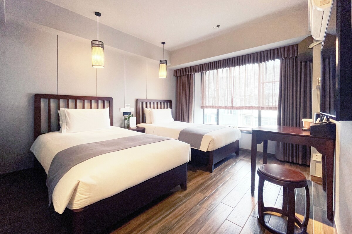Quarto de hotel com duas camas, uma escrivaninha, uma cadeira e uma janela com cortinas.