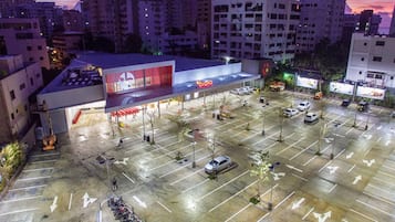 Um estacionamento com vários carros e motocicletas, um edifício grande com a placa 'Bravo' e edifícios altos ao redor.