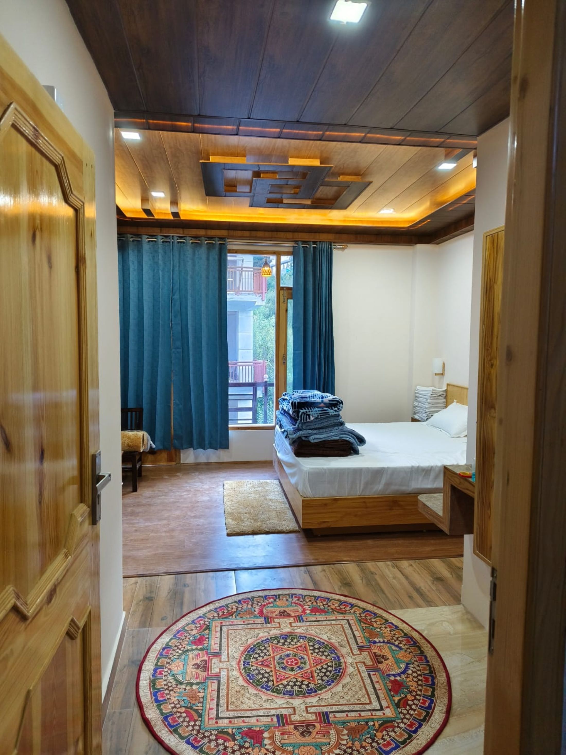 Quarto com cama, janela com cortinas, piso de madeira e tapete estampado.