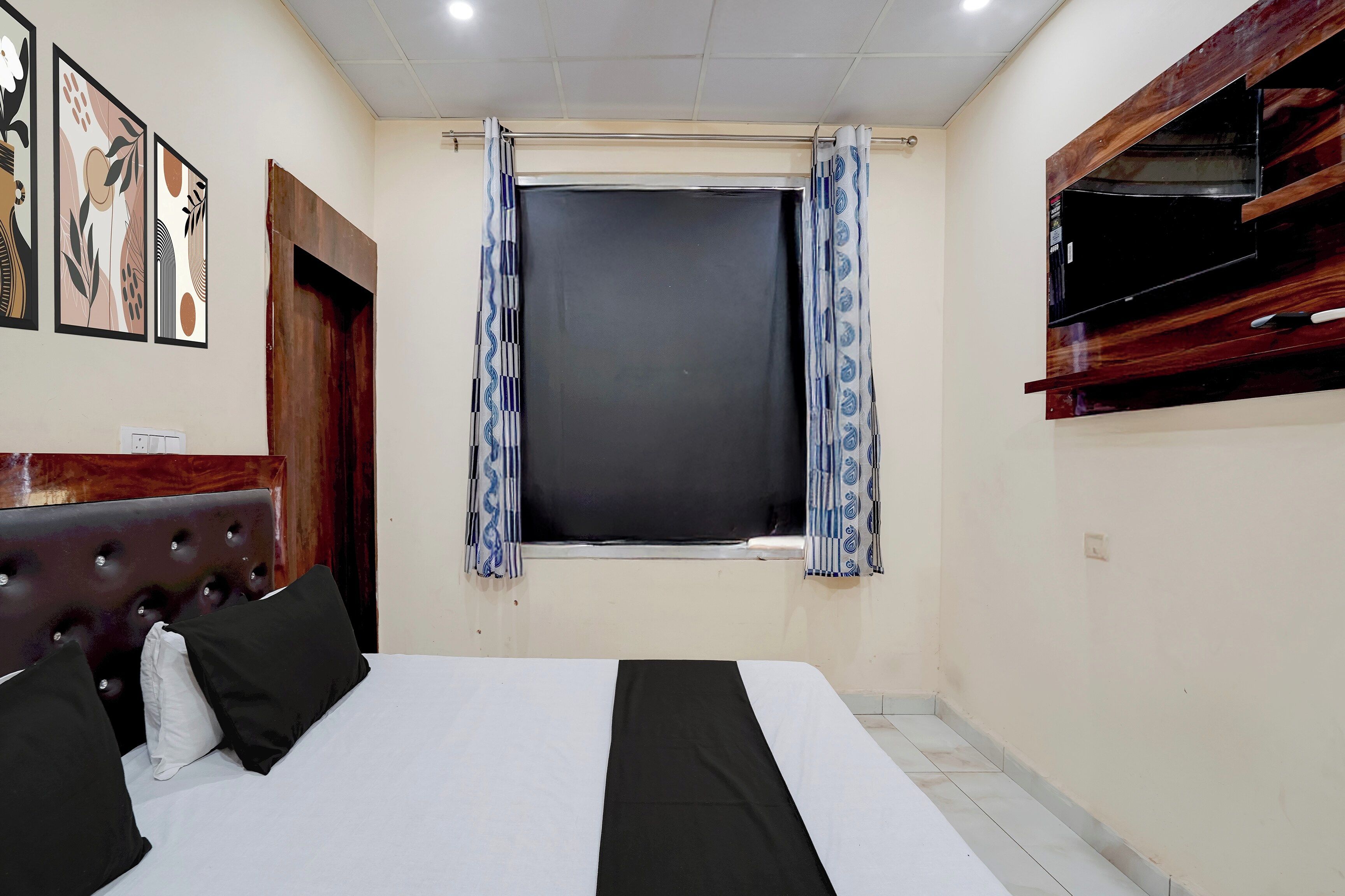 Quarto de hotel com cama, janela com cortinas, televisão fixada na parede e quadro decorativo na parede.
