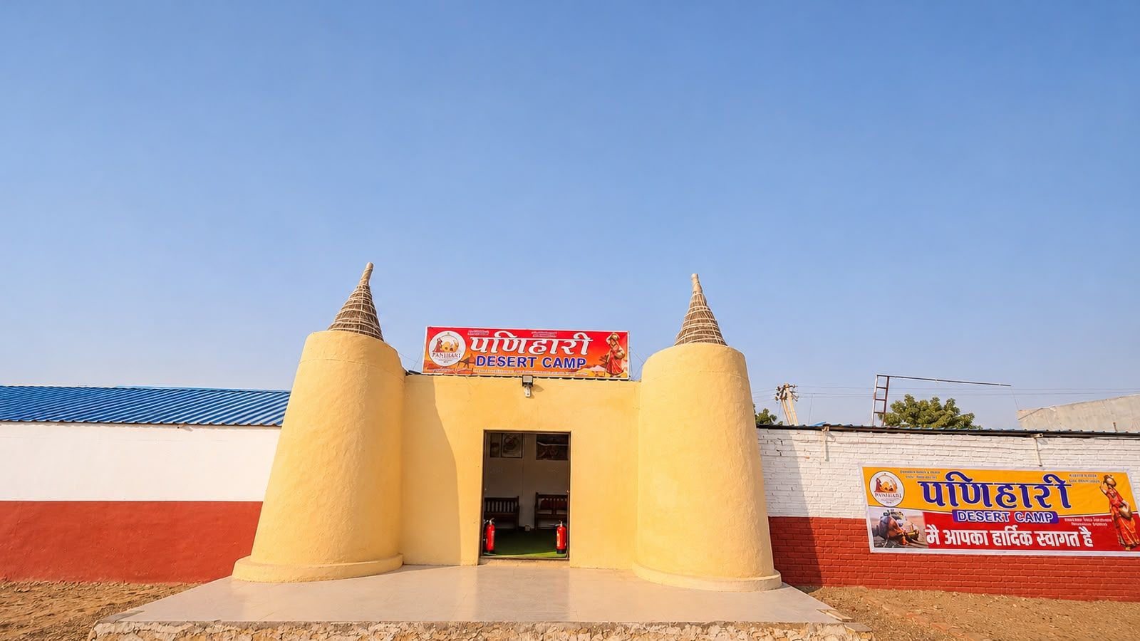 Entrada de um acampamento no deserto, com uma placa e uma parede de tijolos.