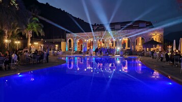 Área de piscina com um edifício de pedra ao fundo, cercada por mesas e cadeiras.