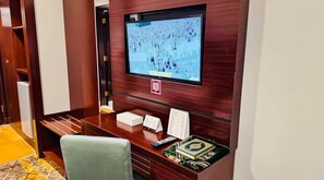 Balcão de recepção de hotel com uma televisão exibindo uma multidão, área de estar e uma pequena mesa com um livro e um controle remoto.