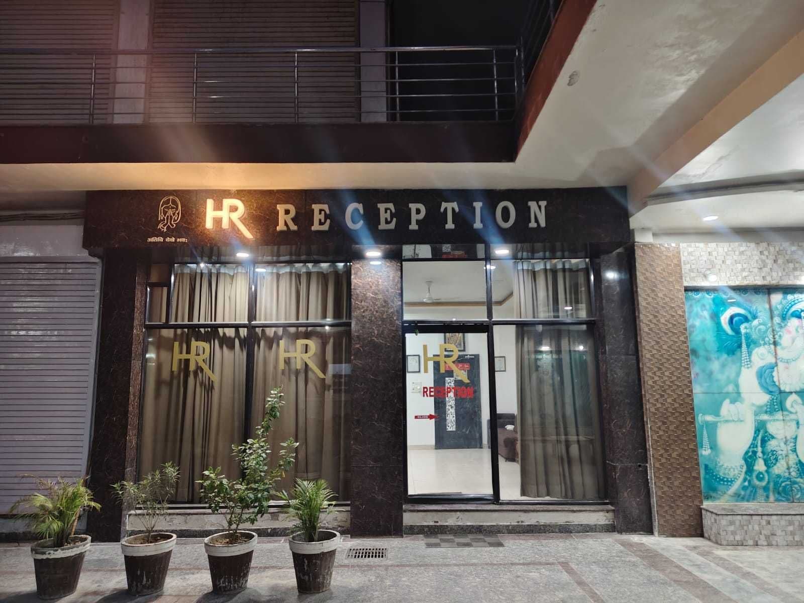 Recepção de hotel com fachada escura, amplas janelas de vidro e plantas em vasos na frente.