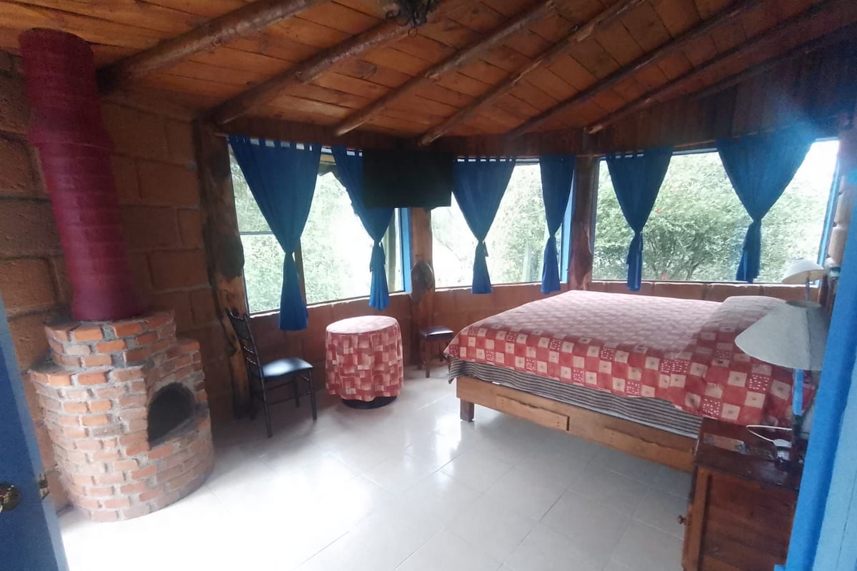 Um quarto com lareira de tijolos, cama com colcha xadrez, mesa redonda e cortinas azuis.