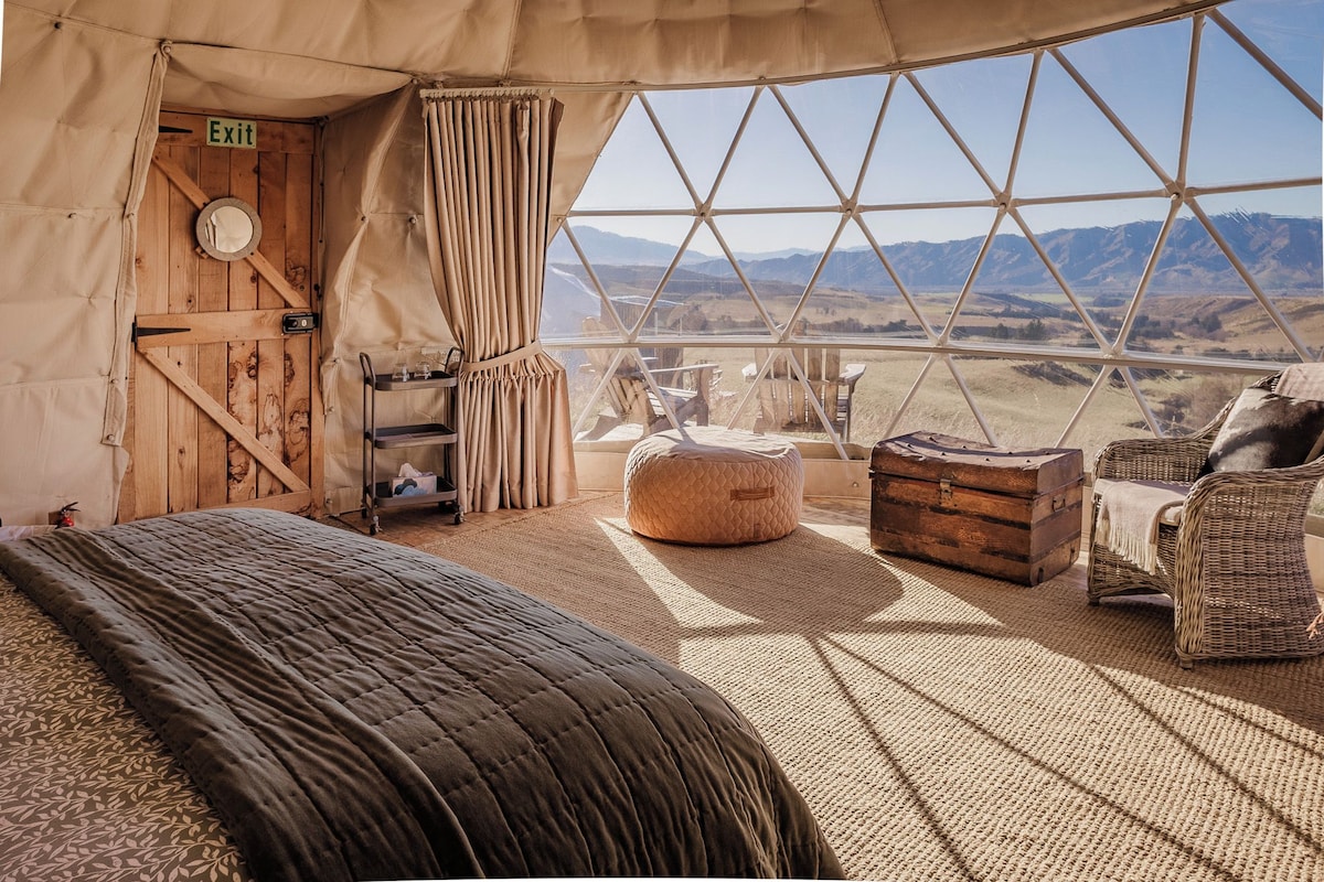 Quarto com tenda, cama, mesa pequena, cadeira e vista para montanhas.
