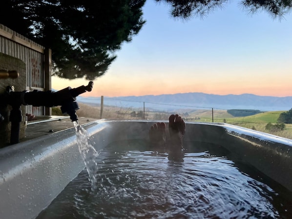 Uma banheira de hidromassagem com uma pessoa dentro, cercada por paisagem natural e montanhas ao fundo.