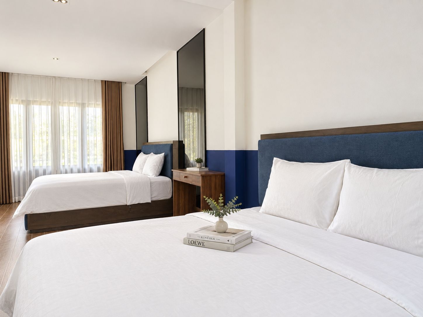 Quarto de hotel com duas camas, uma mesa de cabeceira com abajur, um espelho e uma janela com cortinas.