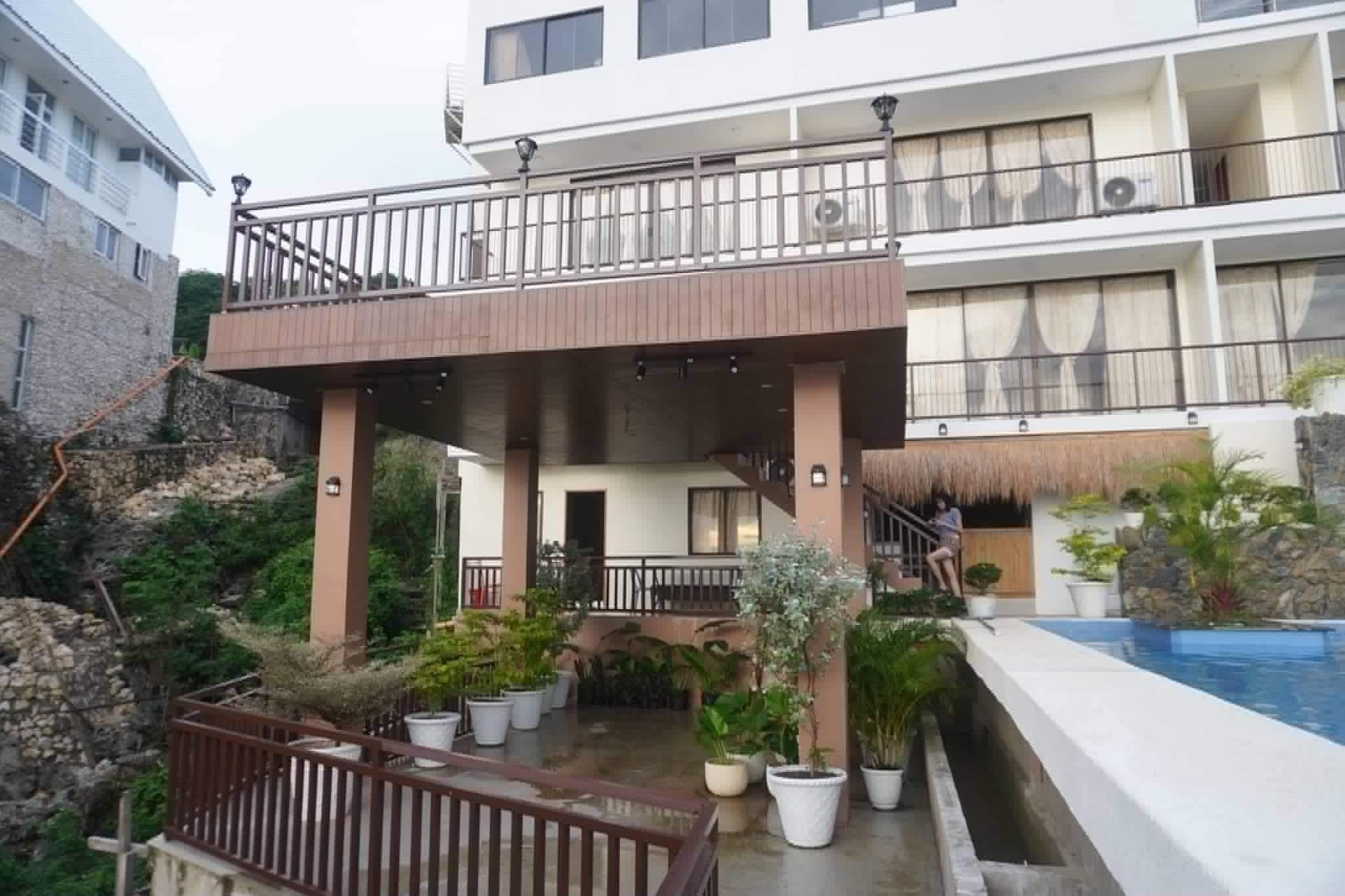 Um edifício com vários andares, área externa coberta, plantas em vasos e piscina.